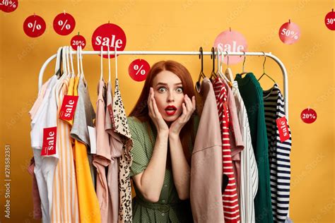 Surprised Girl With Palms On Her Cheek Standing Among Clothes In The Fitting Room Close Up
