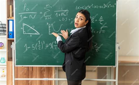 запутанная молодая учительница стоящая перед доской указывает на доску в классе Бесплатно Фото
