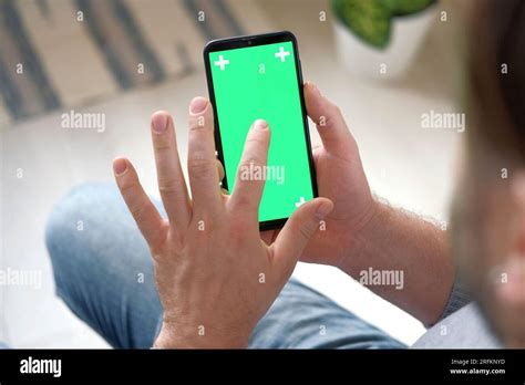 Young Man Sitting At Home Holding Smartphone Green Mock Up Screen In Hand Male Person Using