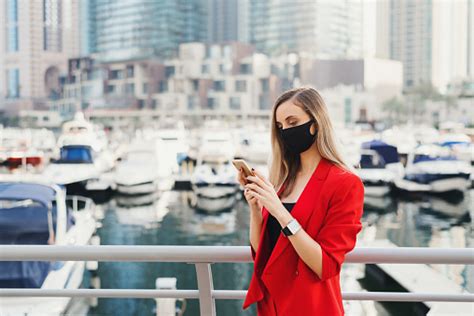 요트와 고층 건물 근처 도심에 서서 스마트 폰 화면을보고 검은 색 보호 얼굴 마스크를 착용한 빨간 양복을 입은 젊은 사업가 새로운 일반 개념 검색에 대한 스톡 사진 및 기타