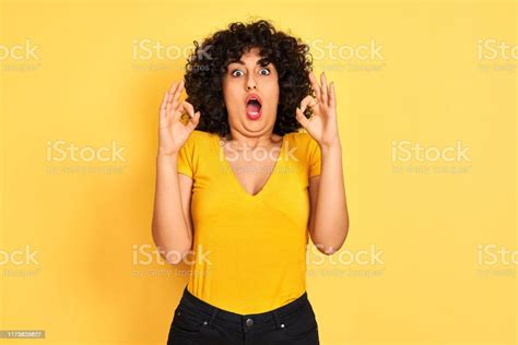 고립 된 노란색 배경 위에 서있는 곱슬 머리를 한 젊은 아랍 여성은 손가락으로 확인 승인 기호를하고 놀랍고 충격을 받았습니다