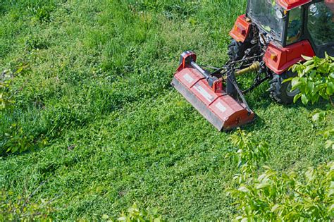 빨간 트랙터에 부착된 잔디 깎는 기계는 잔디를 깔끔하게 자르는데 이는 정밀함 기술 및 농업 노동에 대한 현대적인 접근 방식을 반영합니다 0명에 대한 스톡 사진 및 기타