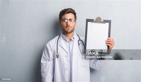 스마트 얼굴 심각한 생각에 자신감이 식 클립보드를 들고 회색 그런 지 벽에 잘생긴 젊은 의사 남자 감정에 대한 스톡 사진 및