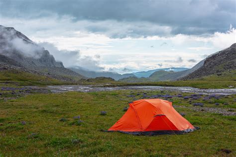 비가 내리는 텐트가 있는 경치 좋은 산의 풍경은 흐린 하늘 아래에서 햇빛에 떨어집니다 비오는 날씨에 녹색 산 계곡에서 보라색 꽃 사이에 잔디에 선명한 오렌지 텐트와 극적인