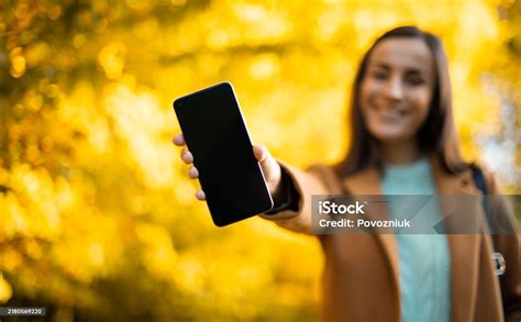 한 젊은 여성이 스마트폰을 카메라를 향해 손을 내밀며 미소를 짓고 있다 휴대폰 화면이 눈에 띄고 배경은 생생한 노란 잎이 있는 흐릿한 가을 풍경입니다 10월에 대한 스톡