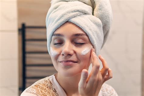 긍정적 인 예쁜 여자의 초상화는 거울에서 데이트 모습을 준비하고 회색 배경 위에 부드러운 신선한 자연을 만들기 위해 그녀의 얼굴에 새로운 스킨 케어 크림을 바릅니다 머리에