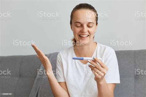 긍정적 인 임신 검사를 들고 피그 테일을 가진 흥분된 젊은 성인 여성은 행복한 낙관적 인 표정으로 결과를 찾고 손을 들어