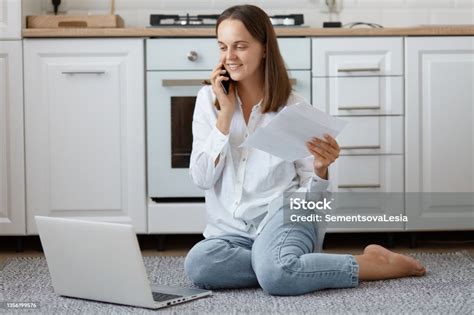 부엌에서 바닥에 앉아 노트북과 스마트 폰을 사용하여 즐거운 대화를 나누고 종이 문서를 들고 있는 매력적인 젊은 성인 여성의 미소 짓는 실내 샷 25 29세에 대한 스톡 사진