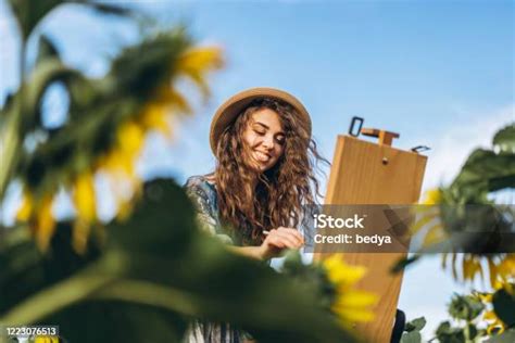 곱슬 머리와 모자를 쓰고 있는 젊은 여성이 자연에 그림을 그리고 있습니다 아름다운 날 해바라기 밭에 서 있는 한 여성 개념에 대한 스톡 사진 및 기타 이미지 Istock