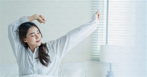 잠옷을 입은 아시아 아름다운 소녀는 아침에 행복으로 일어난다 매력적인 젊은 여성은 웃고 행복하고 긴장을 풀고 집 침실의 침대에서 잠에서 깨어난 후 몸을 스트레칭합니다