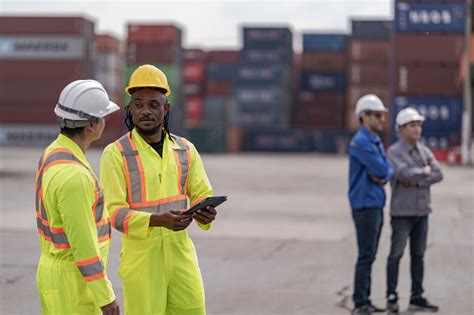 눈에 잘 띄는 안전 장비와 안전모를 쓴 두 명의 물류 작업자가 바쁜 선적 컨테이너 야적장에서 작업에 대해 논의하고 있습니다 한 명은 태블릿을 들고 화물 더미를 가리키고