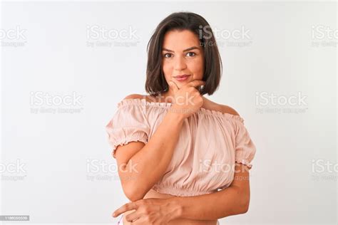 고립된 흰색 배경 위에 서있는 캐주얼 한 티셔츠를 입은 젊은 아름다운 여성은 턱에 손을 들고 교차 된 팔과 손으로 미소 짓는 카메라를 보고 자신감을 보입니다 긍정적 인 생각