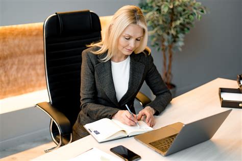 성공적이고 영향력 있는 성숙한 집중 자신감 있는 백인 금발 사업가 브로커 또는 금융 전문가 노트북과 함께 사무실 테이블에 앉아 메모를 하고 비즈니스 전략을 개발합니다 45