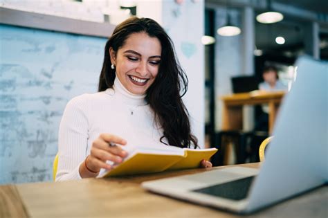 대학 시험까지 준비하는 지식 교과서에서 노트를 읽는 갈색 머리 머리 쾌활한 여성 탐색을위한 노트북 장치로 테이블 근처에서 종이 정보를 공부하는 행복한 여학생 20 24세에
