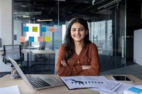 차트와 노트북이 있는 현대적인 사무실의 책상에 앉아 팔짱을 낀 자신감 넘치는 라틴 아메리카 사업가가 성공과 리더십을 반영합니다 경영자에 대한 스톡 사진 및 기타 이미지