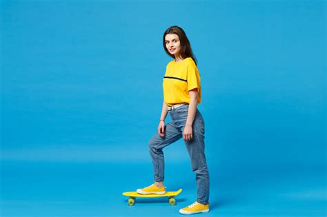 스튜디오에서 파란색 벽 배경에 고립 된 노란 스케이트 보드와 서 카메라를 찾고 생생한 캐주얼 복장에 매력적인 젊은 여자의 측면 보기 사람들 라이프 스타일 개념입니다 복사
