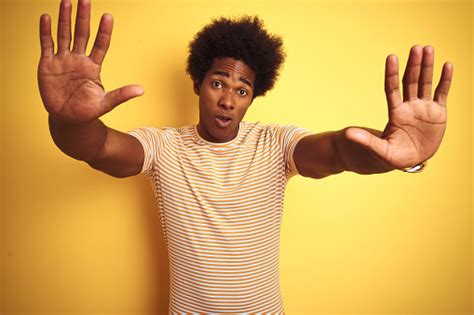 고립 된 노란색 배경 위에 서있는 줄무늬 티셔츠를 입은 미국 남자가 손바닥 화가 나고 좌절하는 표정으로 중지 제스처를하고 있습니다 T 셔츠에 대한 스톡 사진 및 기타 이미지