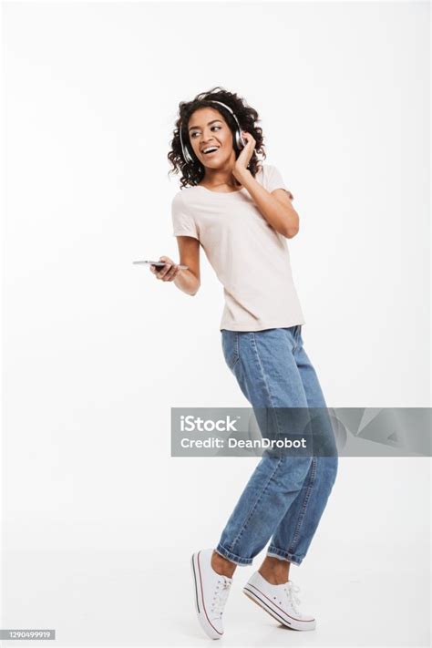 흰색 배경에 고립 된 무선 헤드폰과 스마트 폰을 사용하여 음악을 듣고 갈색 곱슬 머리를 가진 귀여운 아프리카 계 미국인 여성의 전체 길이 이미지 갈색에 대한 스톡 사진 및