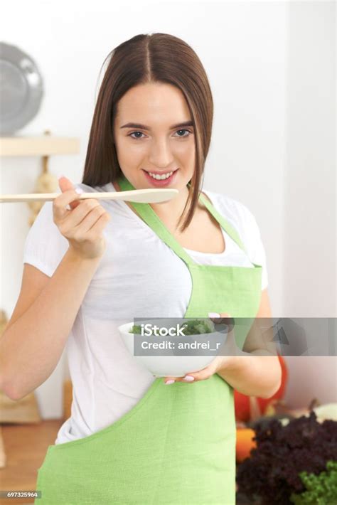 녹색 앞치마에 젊은 갈색 머리 여자 요리 또는 부엌에서 신선한 샐러드를 먹는 나무 숟가락과 그릇에 그녀의 손 잡고 주부 음식과 건강 개념입니다 가정 생활에 대한 스톡 사진