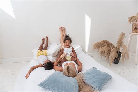 젊은 백인 금발의 어머니가 놀고 아프리카 계 미국인 딸 아들과 함께 흰색 침대에 누워 있습니다 행복한 다민족 가족의 아침 여자는 포옹 부드러움으로 아이들을 스트로크 미니멀한