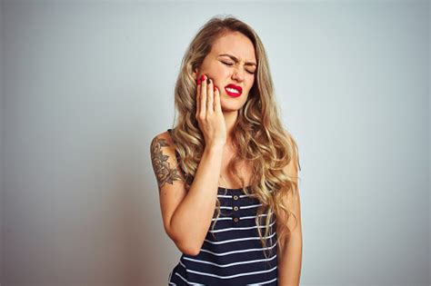 치통이나 치아 질환으로 인해 고통스러운 표정으로 입을 만지는 흰색 고립 된 배경 위에 서있는 줄무늬 티셔츠를 입은 젊은 아름다운 여성 치과 의사 개념 T 셔츠에 대한 스톡