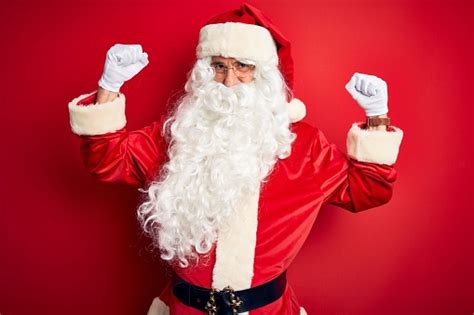 고립된 빨간 배경 위에 서있는 산타 의상을 입은 중년 잘생긴 남자가 자랑스럽게 웃고있는 팔 근육을 보여줍니다 피트니스 개념입니다 12월에 대한 스톡 사진 및 기타 이미지