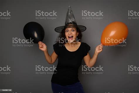 마법사 모자를 쓰고 행복한 즐거운 아름다운 히스패닉 여성이 주황색과 검은 색의 공중볼 두 개를 들고 복사 공간이있는 회색 배경에 포즈를 취하는 카메라를 봅니다 할로윈 컨셉