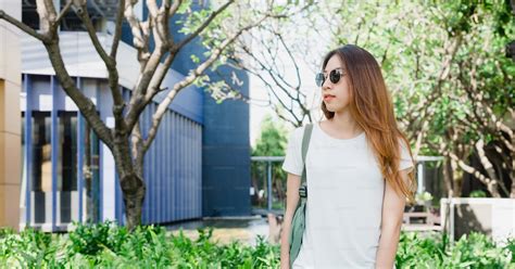 흰색 빈 티셔츠를 입은 아시아 힙스터 소녀 긴 갈색 머리가 길 한가운데에 서 있다 스트리트웨어를 입은 여성이 녹색 도시 배경에 서 있습니다 텍스트 또는 디자인을 위한