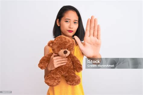 진지하고 자신감있는 표정 방어 제스처로 정지 표지판을하고 열린 손으로 고립 된 흰색 배경 위에 서있는 테디 베어를 들고있는 젊은 아름다운 중국 여성 경계 표지에 대한 스톡