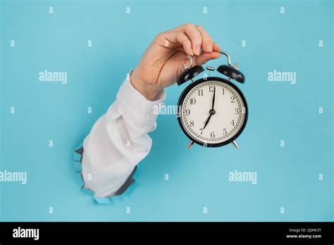 A Womans Hand Sticks Out Of A Hole In A Blue Paper Background And Holds An Alarm Clock Stock