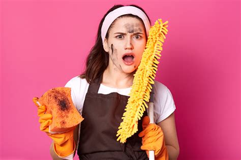 흰색 캐주얼 T 셔츠 갈색 앞치마와 헤어 밴드 들고 스폰지와 노란 걸 레 충격에 놀란 유럽 젊은 Housewoman는 매우 더러운 방을 청소 해야 합니다 분홍색 스튜디오