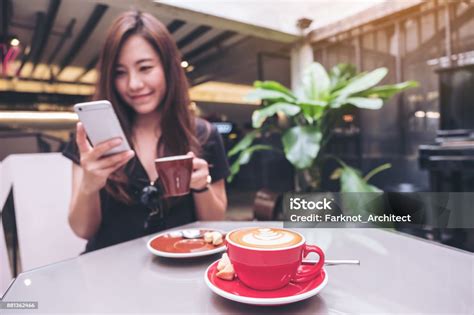 들고 고 다락방 카페에서 라 떼 커피 컵 유리 테이블에 커피를 마시는 동안 스마트폰 사용 하 여 아름 다운 아시아 여자의 근접 촬영 이미지 라떼에 대한 스톡 사진 및 기타