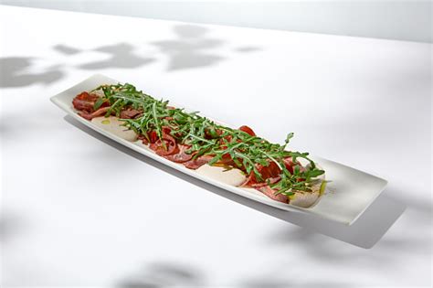 가혹한 그림자가있는 흰색 테이블에 Vitello Tonnato 참치 소스와 Arugula와 함께 얇게 썬 송아지 고기 여름날 세라믹 접시에 Vitello Tonnato 미적