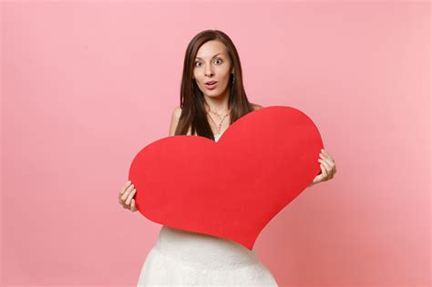 아름 다운 하얀 웨딩에서 충격된 놀된 신부 여자의 초상화 드레스 파스텔 핑크 배경에 고립 된 빈 빈 빨간 심 혼을 들고 결혼식 축 하 개념입니다 광고에 대 한 공간을 복사