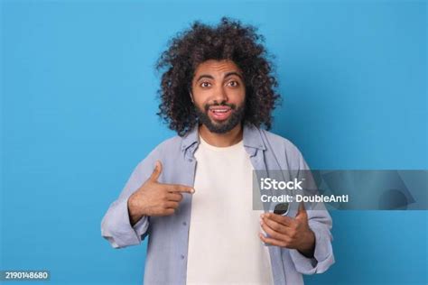 곱슬 머리를 가진 매력적인 젊은 남자는 생생한 파란색 환경에서 스마트 폰을 보여줍니다 30 34세에 대한 스톡 사진 및 기타 이미지 30 34세 Brand Name