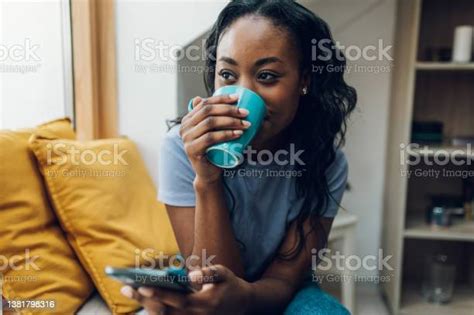 커피 한 잔과 스마트폰으로 창턱에 앉아 있는 아프리카계 미국인 여성 20 24세에 대한 스톡 사진 및 기타 이미지 20 24세 Brand Name Smart Phone