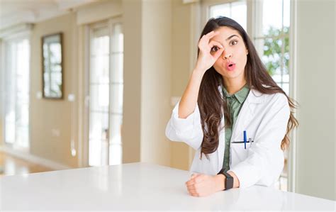 치료 치료사 또는 의사로 병원에서 의료 코트를 착용 하는 젊은 여성은 놀란 얼굴 눈을 통해 보고 충격을 확인 제스처를 하 고