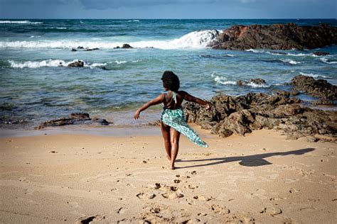 검은 파워 머리를 가진 아름다운 여자가 긴 드레스를 들고 해변 모래사장을 걷고 있다 배경에는 하늘 구름 바위 바다가 있습니다 30 34세에 대한 스톡 사진 및 기타 이미지
