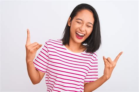 스트라이프 티셔츠를 입은 젊은 중국 여성이 고립된 흰색 배경 위에 서서 미친 표정으로 손을 들고 바위 기호를 하고 있습니다 음악 스타 무거운 개념 T 셔츠에 대한 스톡 사진