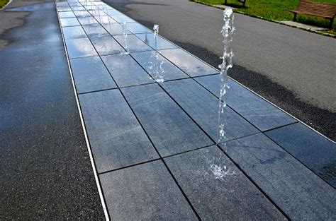 보도의 스트립에 쌓인 사각형 화강암 포장 실린치 번갈아 스프레이 높이가있는 물 스프레이 물 특징은 공원 줄의 자갈이 깔린 광장에서 직접 나타납니다 산책로의 젖은 타일 젖은에
