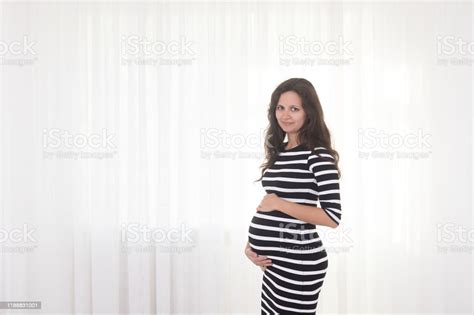 줄무늬 드레스를 입은 임산부는 흰색 배경에 배에 손을 대고 있습니다 임신 출산 준비 및 기대 개념 임신의 아름다운 부드러운 분위기 사진 갈색 머리에 대한 스톡 사진 및 기타