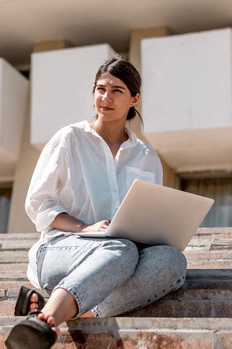 하얀 셔츠와 청바지를 입은 젊은 검은 머리 여성은 여름날 도시 거리의 계단에 앉아있는 노트북을 사용합니다 원격 근무 프리랜서 온라인 교육 갈색 머리에 대한 스톡 사진 및