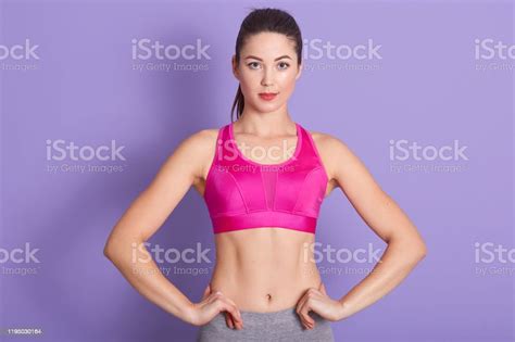 분홍색 운동 복장과 포니 테일을 가진 Winsome 백인 여성 라일락 배경 심각한 스포티 한 여성 케핑 손을 엉덩이에 놓고 카메라를 직접 보고 있는 포즈를 취합니다 피트니스