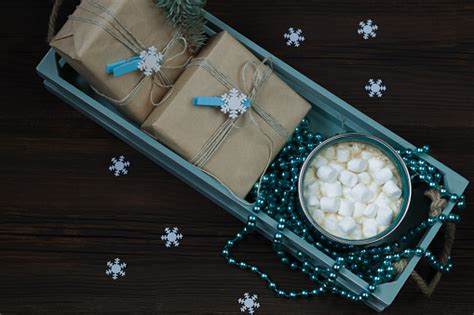 크래프트 페이퍼 눈송이 마시멜로가 달린 따뜻한 음료로 장식된 크리스마스 선물 상자는 어두운 배경에 구슬이 있는 청록색 나무 상자에 들어 있습니다 겨울 설날 테마 12월에