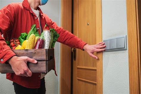 현관에 음식과 종이 봉투를 들고 보호 마스크에 배달 남자 택배는 문에 서서 고객의 초인종을 울립니다 그는 신선한 채소와 과일 한