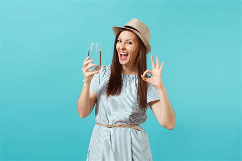 블루 드레스 모자 들고와 유리 파란색 배경에 고립에서 분명 신선한 순수한 물을 마시는 젊은 아름 다운 여자의 초상화 건강 한 생활양식 사람들이 진심으 감정 개념입니다 공간에