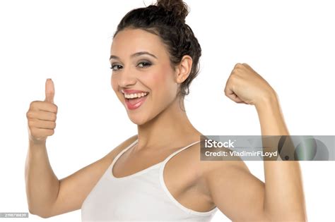 흰 탱크 탑을 입고 롤빵에 검은 머리를 한 열정적인 여자 엄지손가락을 치켜세우고 팔 근육을 구부립니다 밝은 미소 흰색 배경에 스튜디오에서 촬영 20 24세에 대한 스톡 사진