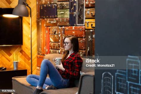 아름 다운 젊은 여성 캐주얼 의류와 안경 음료 커피 현대적인 인테리어의 배경에서 소파에 앉아 벽의 오래 된 가방 노트북에 대한