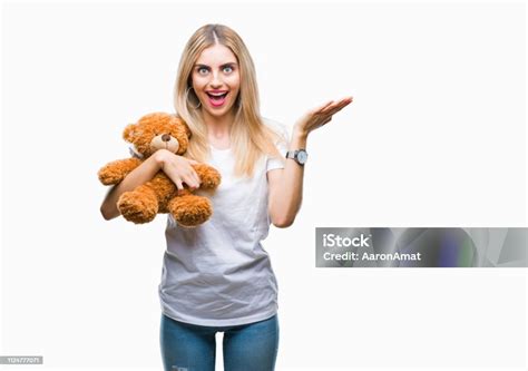 젊은 아름 다운 금발 여 자가 고립 된 배경 위에 테 디 베어를 들고 매우 행복 하 고 흥분 승자 표현은 큰 웃음과 손을 제기와 비명을 축 하 귀여운에 대한 스톡 사진 및