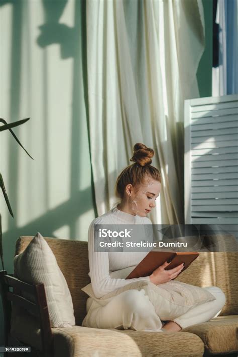 소파에 앉아 집에서 미용 절차를 하는 동안 책을 읽으면서 즐거운 시간을 보내는 아름다운 아가씨 소녀 얼굴 치료 테라피 트리트먼트 뷰티 스킨 케어 마스크 팩에 대한 스톡 사진